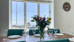 Uma mesa de jantar posta com pratos, copos e um vaso de flores, com vista para edifícios através de amplas janelas.