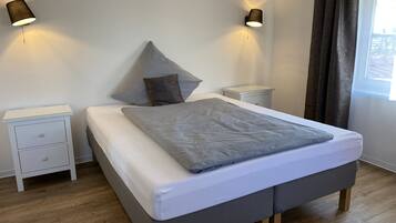 Quarto com cama, duas mesas de cabeceira, um criado-mudo, janela com cortinas e iluminação na parede.