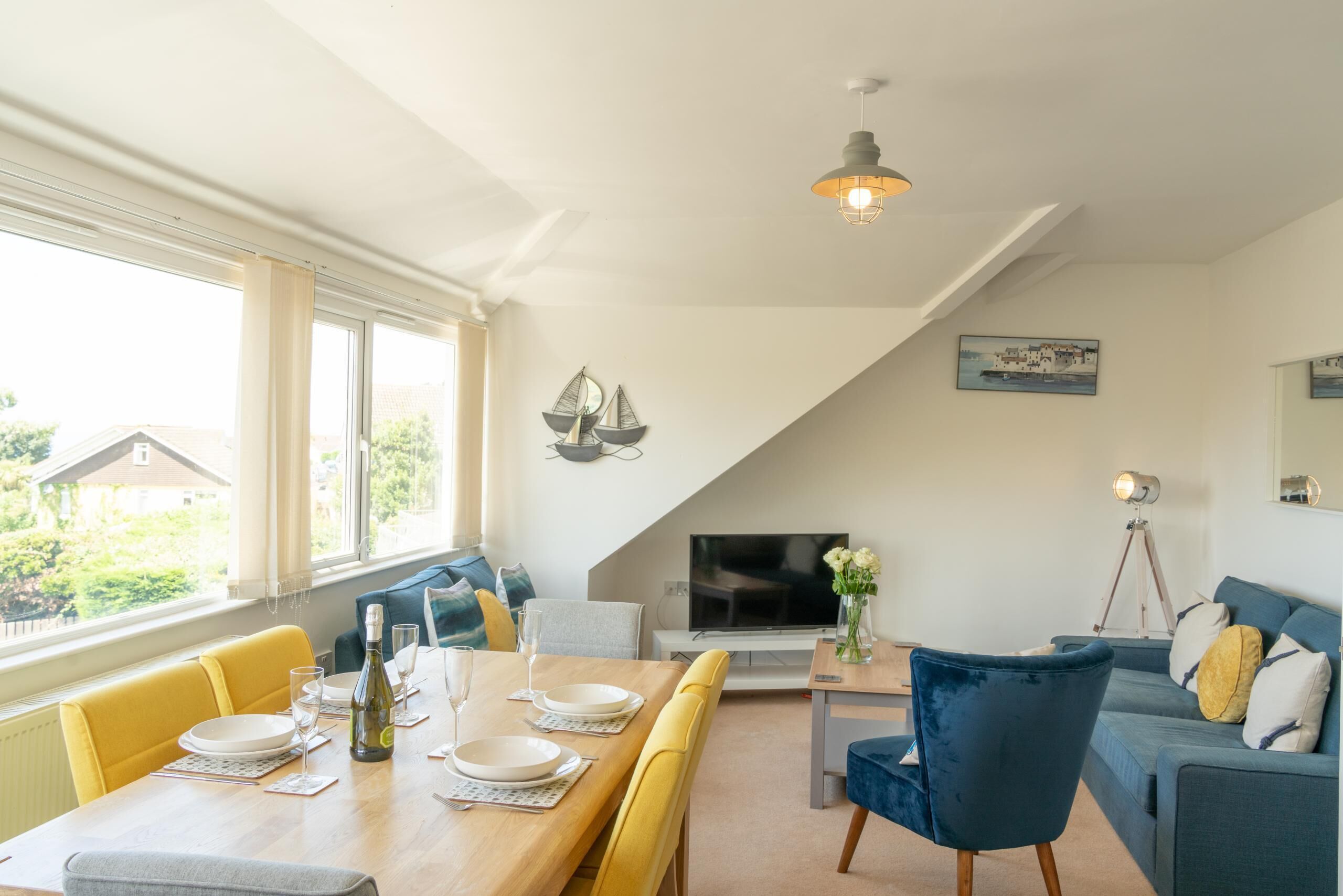 Uma sala de jantar com uma mesa de madeira posta para quatro pessoas, um sofá e uma televisão.
