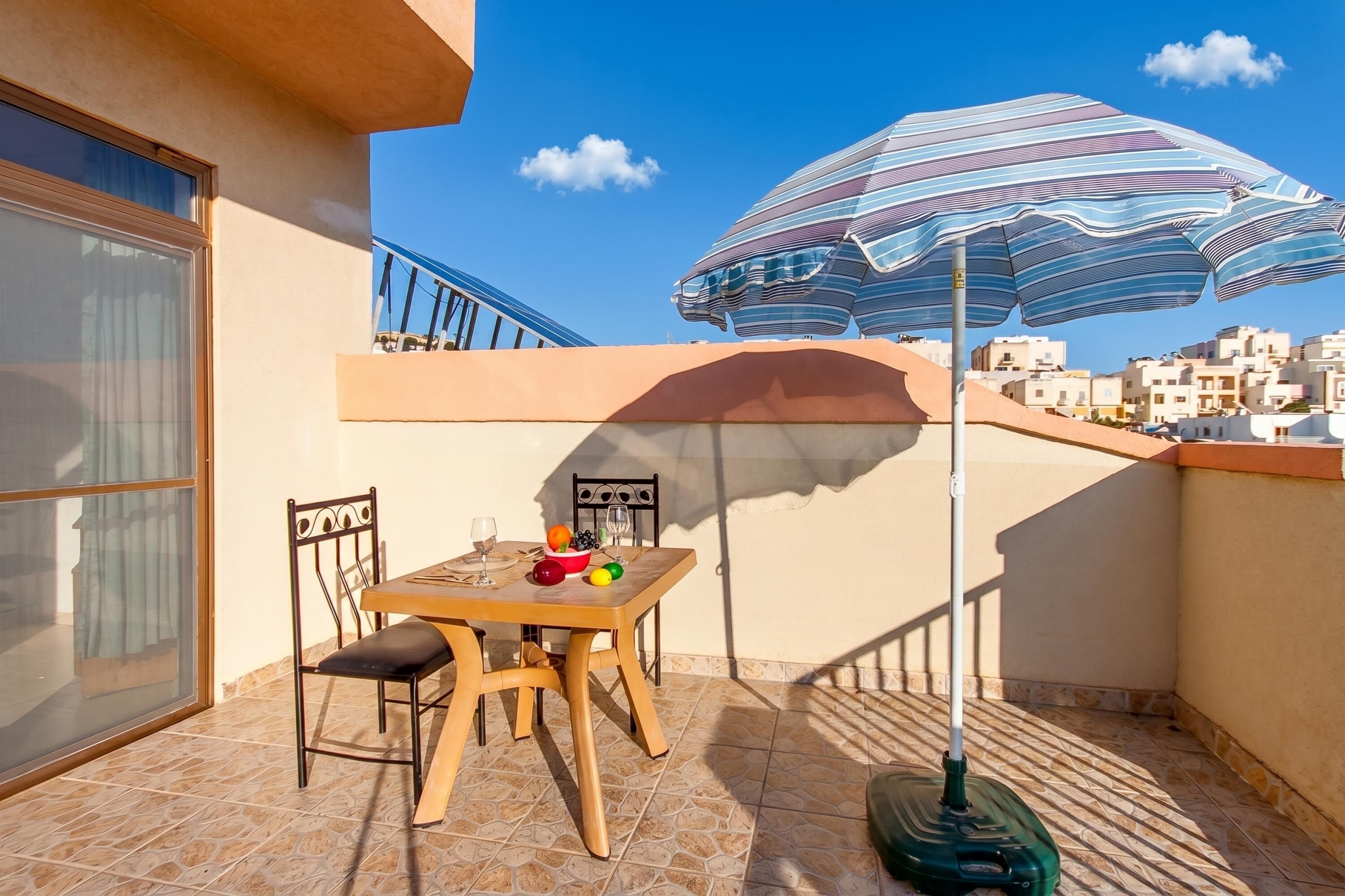 Uma varanda com mesa, cadeiras, um guarda-sol listrado e uma mala.