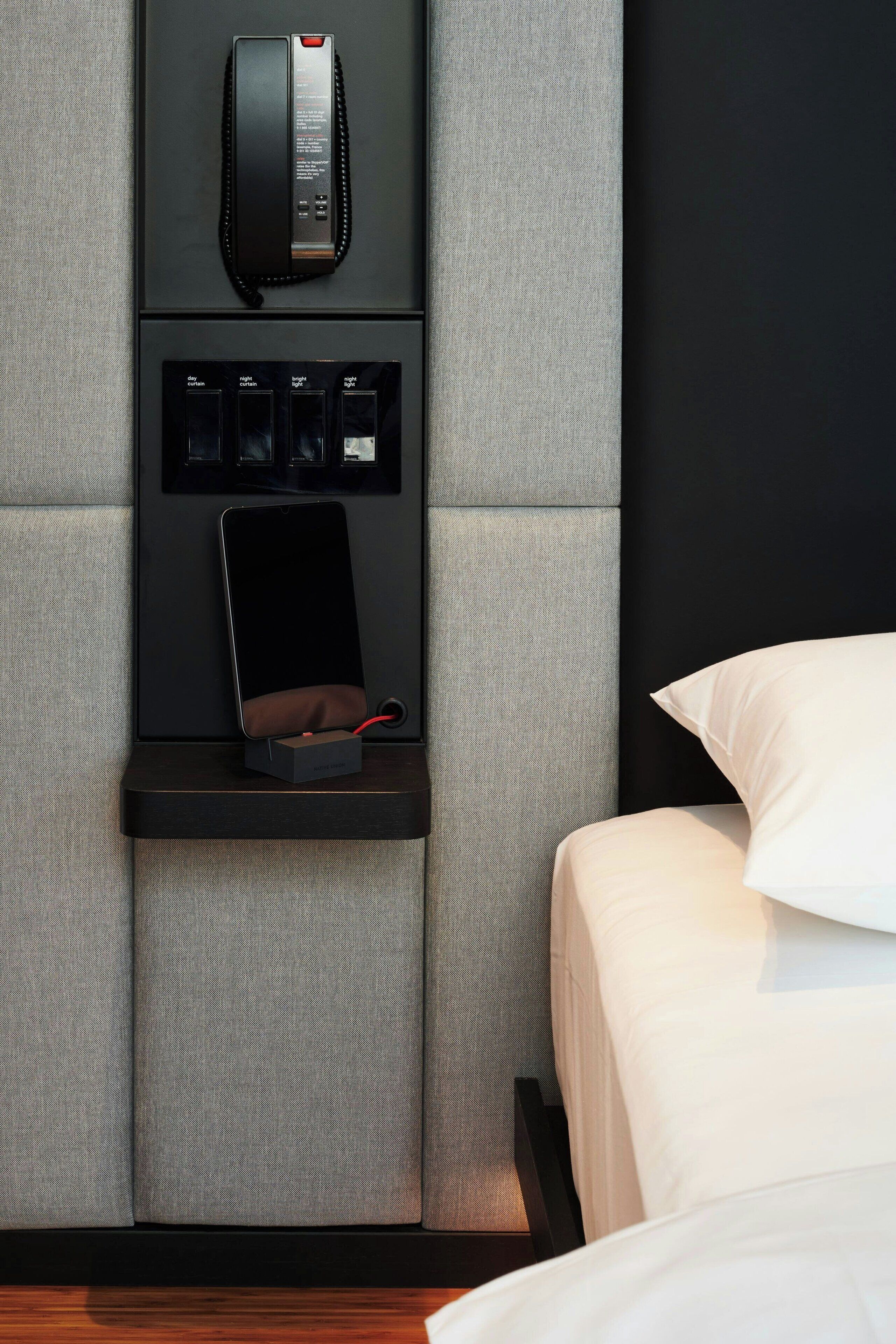 Quarto de hotel com cama, mesa de cabeceira com ponto de carregamento para celular e painel de controle fixado na parede.