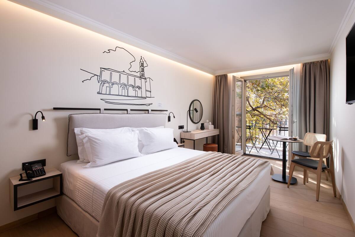 Quarto de hotel moderno com uma cama grande, uma mesinha de cabeceira com telefone, uma pequena mesa com uma cadeira e uma varanda com vista para árvores e edifícios.