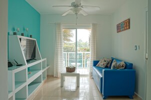 Uma sala de estar com um sofá azul, um rack branco para TV, um ventilador de teto e vista para o exterior por uma porta de vidro.