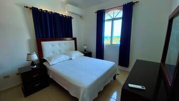 Quarto de hotel com cama, mesas de cabeceira, abajur, televisão e janela com cortinas.