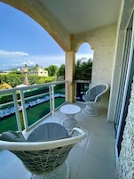 Varanda com duas cadeiras de vime e uma mesa pequena, com vista para um jardim com um espelho d'água.