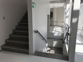 Uma escada com corrimão, um sinal de saída de emergência verde e uma parede de cor clara.