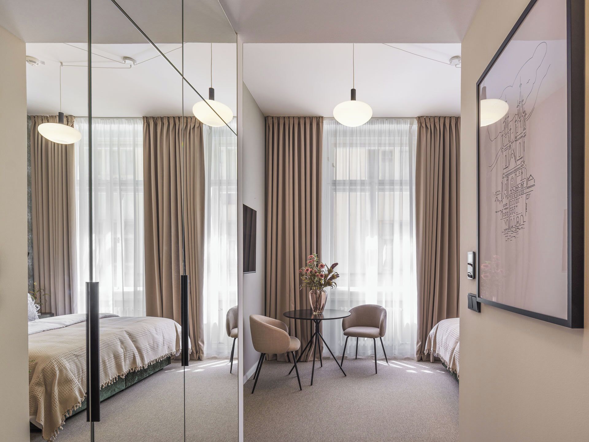 Quarto de hotel moderno com uma cama, duas cadeiras, uma mesinha e um espelho grande.