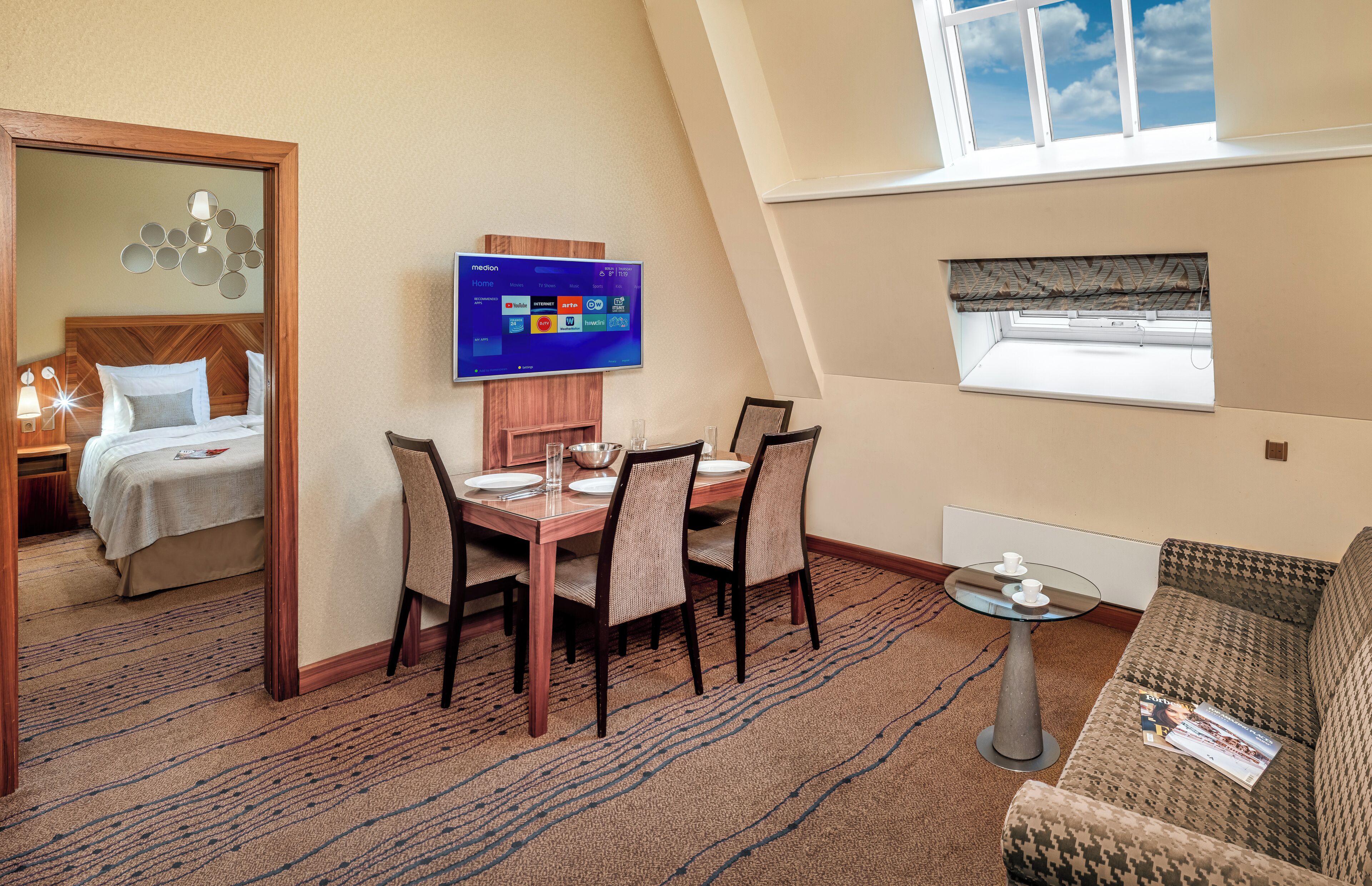 Quarto de hotel com área de refeições, sofá, televisão e janela.