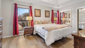 Quarto com cama, cómoda de madeira, mesas de cabeceira, ventoinha no teto e uma janela grande com cortinas vermelhas.