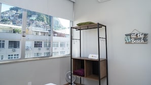 Quarto com uma janela grande, uma estante de metal, um ventilador e uma placa na parede.