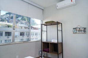 Quarto com uma janela grande, uma estante de metal, um ventilador e uma placa na parede.