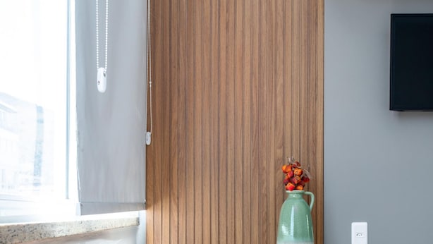 Quarto moderno com parede de ripas de madeira, uma cadeira preta, uma mesinha com um vaso e uma TV fixada na parede.