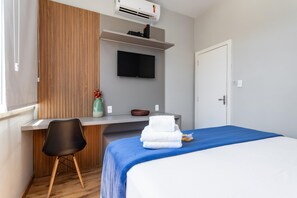 Quarto de hotel com cama, escrivaninha com cadeira, TV de tela plana e ar condicionado.