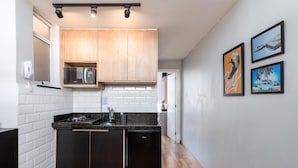Cozinha moderna com armários pretos, bancada preta e micro-ondas embutido. Há armários de madeira acima do micro-ondas e uma geladeira à direita. A cozinha possui azulejos tipo metrô brancos e piso de madeira.