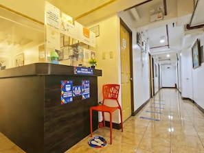 Uma área de recepção de hotel com balcão, cartazes informativos, uma cadeira vermelha e piso revestido de azulejos.