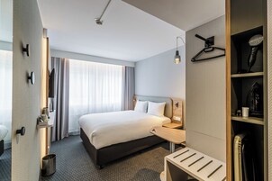 Quarto de hotel moderno com uma cama grande, mesa de cabeceira e escrivaninha com cadeira. Há uma janela com cortinas, luminária na parede e um cabideiro com um casaco.
