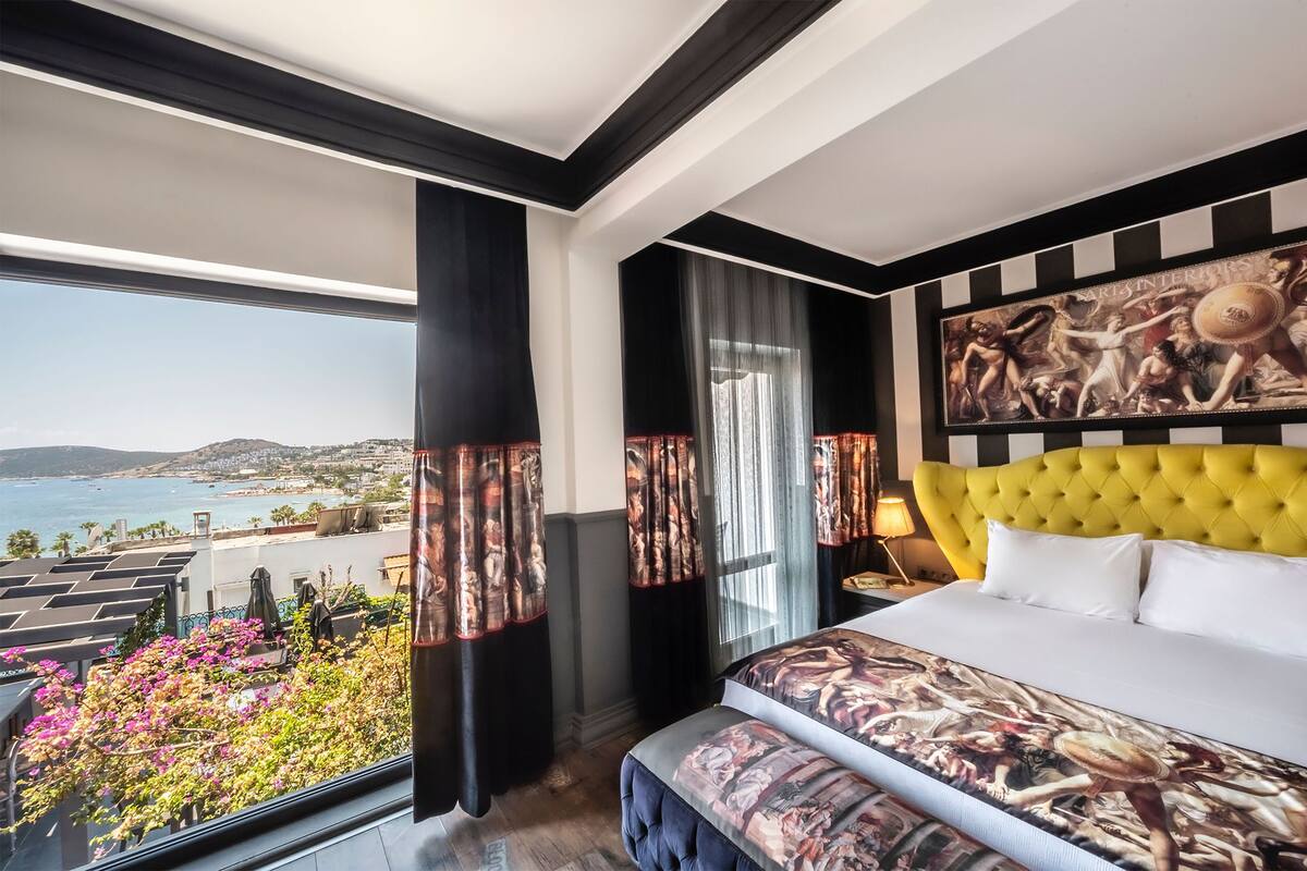 Quarto de hotel com uma cama grande, cabeceira amarela com detalhes embutidos, vista para o mar e varanda florida.