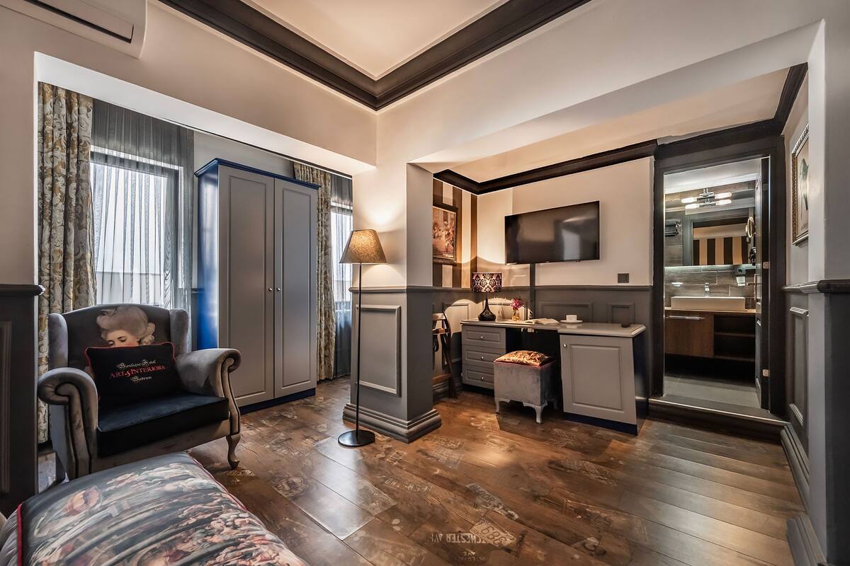 Quarto de hotel compacto com piso de madeira escura, sofá, cadeira, escrivaninha com luminária, TV fixada na parede e vista para o banheiro através de uma porta de vidro.