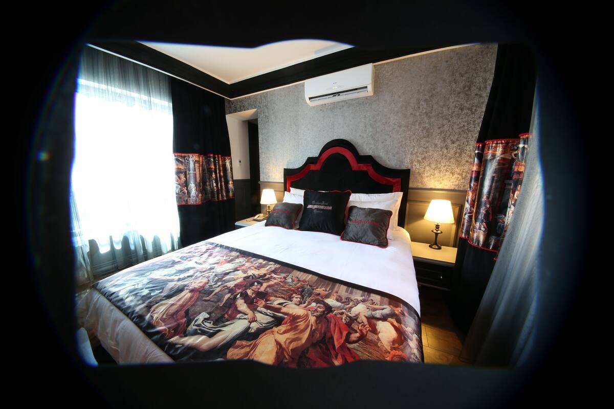 Quarto de hotel com uma cama grande, cabeceira, duas mesas de cabeceira com luminárias e uma janela com cortinas.