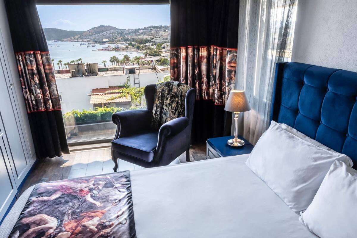 Quarto de hotel com cama, cadeira, mesa de cabeceira e vista para o mar e edifícios.