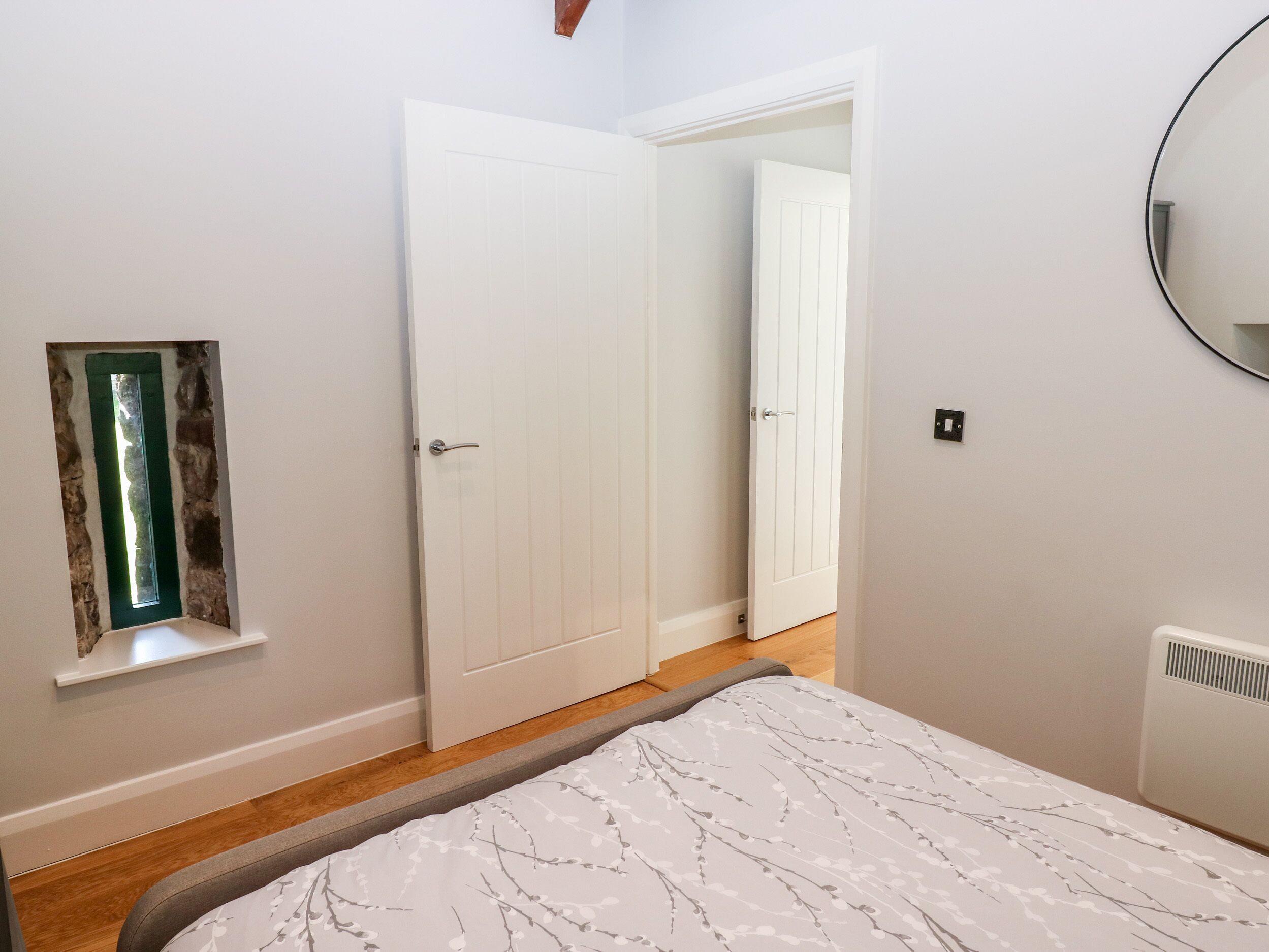 Quarto com uma cama, porta branca, janela pequena e espelho na parede.