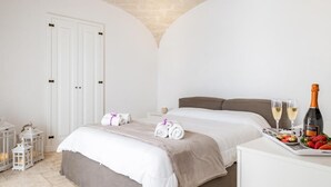 Quarto de hotel com cama, mesa de cabeceira com garrafa e copos, e bandeja com comida e fruta.
