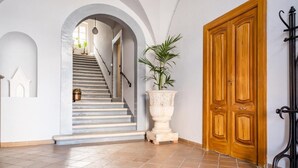 Um corredor com piso de azulejos, escada, porta de madeira e um vaso de plantas.