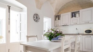 Uma sala de jantar clara com uma mesa, cadeiras e um arranjo de flores. Há uma cozinha com armários brancos e micro-ondas.