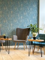 Uma área de estar moderna com parede azul texturizada, uma poltrona cinza com capitonê, um sofá verde e duas mesas de madeira com plantas em vasos.