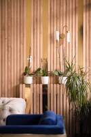 Quarto com parede de ripas de madeira, três plantas em vasos, um sofá azul e luminária fixada na parede.