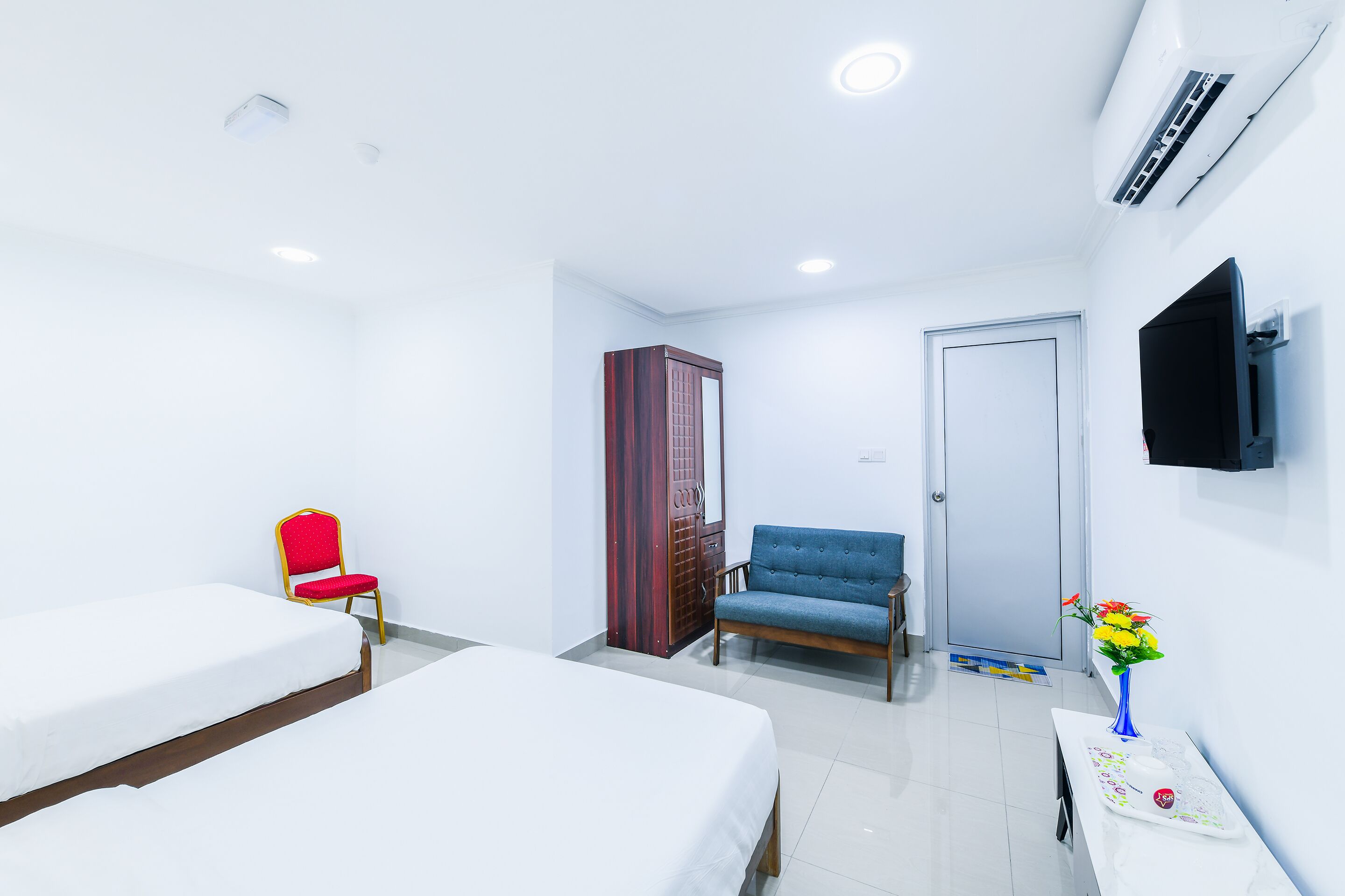Quarto de hotel com duas camas, uma cadeira vermelha, um sofá azul, um guarda-roupa de madeira, uma televisão na parede e uma mesinha com um vaso de flores.