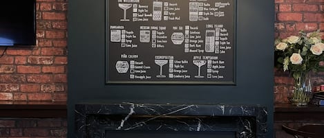 Uma área de bar com um quadro-negro listando coquetéis, cadeiras de couro e uma lareira.