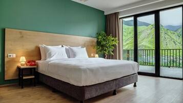 Quarto com cama grande, mesa de cabeceira, luminária, planta e vista para colinas verdes através de uma varanda.