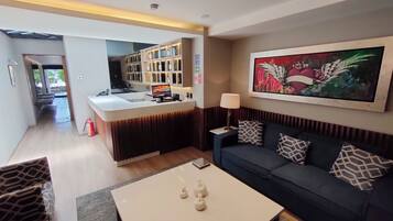 Quarto de hotel moderno com um sofá escuro, uma mesa de centro branca e uma pintura na parede. Há uma receção com um computador e um telefone.
