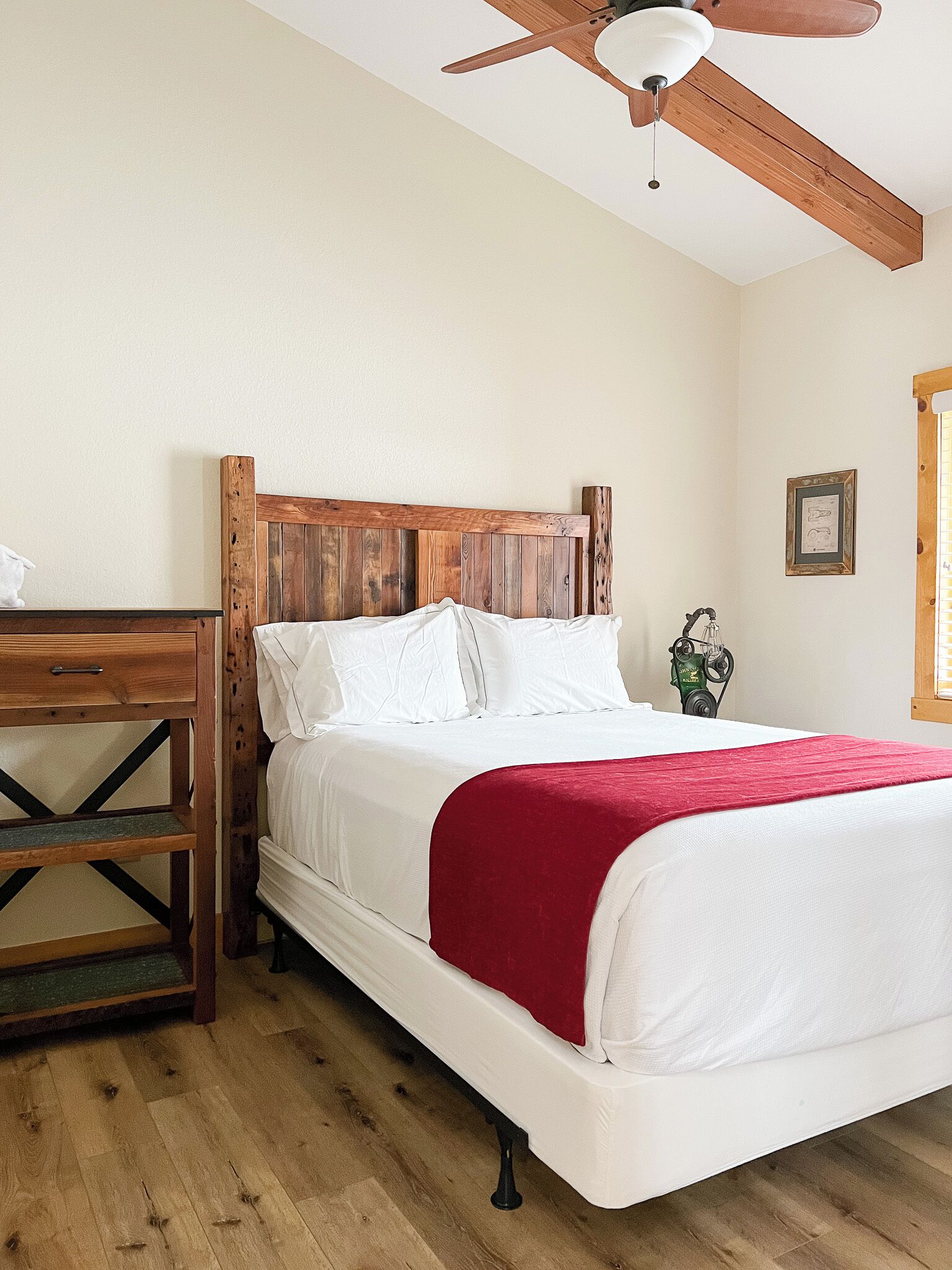 Quarto com cabeceira de madeira, cama com roupa de cama branca e um cobertor vermelho, mesa de cabeceira de madeira e um ventilador de teto.