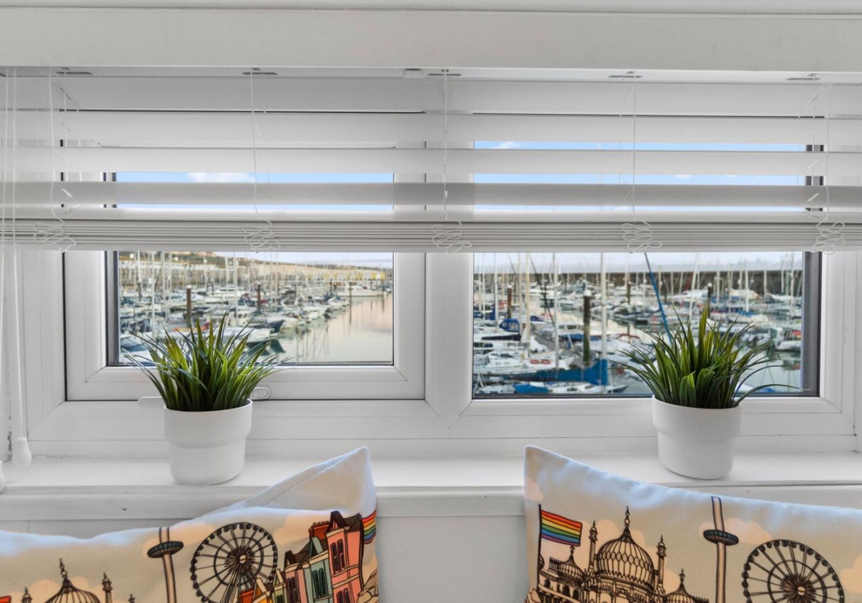 Janela com duas plantas em vasos e dois travesseiros estampados com vista urbana.