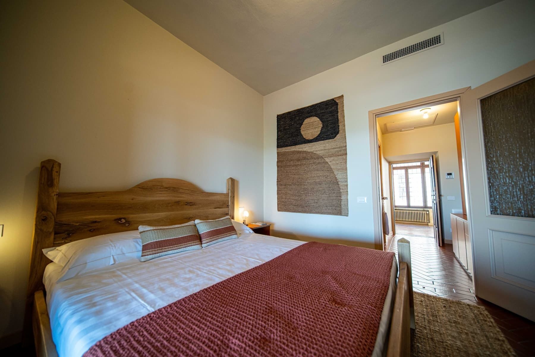 Quarto com cabeceira de madeira, cama com roupa de cama branca e um cobertor vermelho, além de uma obra de arte na parede.