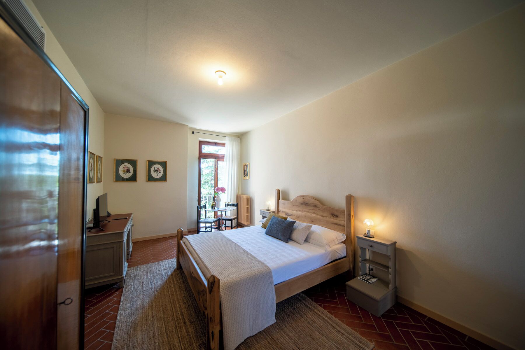 Quarto com cama, mesa de cabeceira e escrivaninha com cadeira. Há uma janela com cortinas e uma porta que dá para outro cômodo.