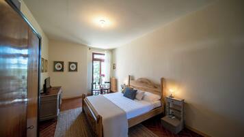 Quarto com cama, mesa de cabeceira e escrivaninha com cadeira. Há uma janela com cortinas e uma porta que dá para outro cômodo.
