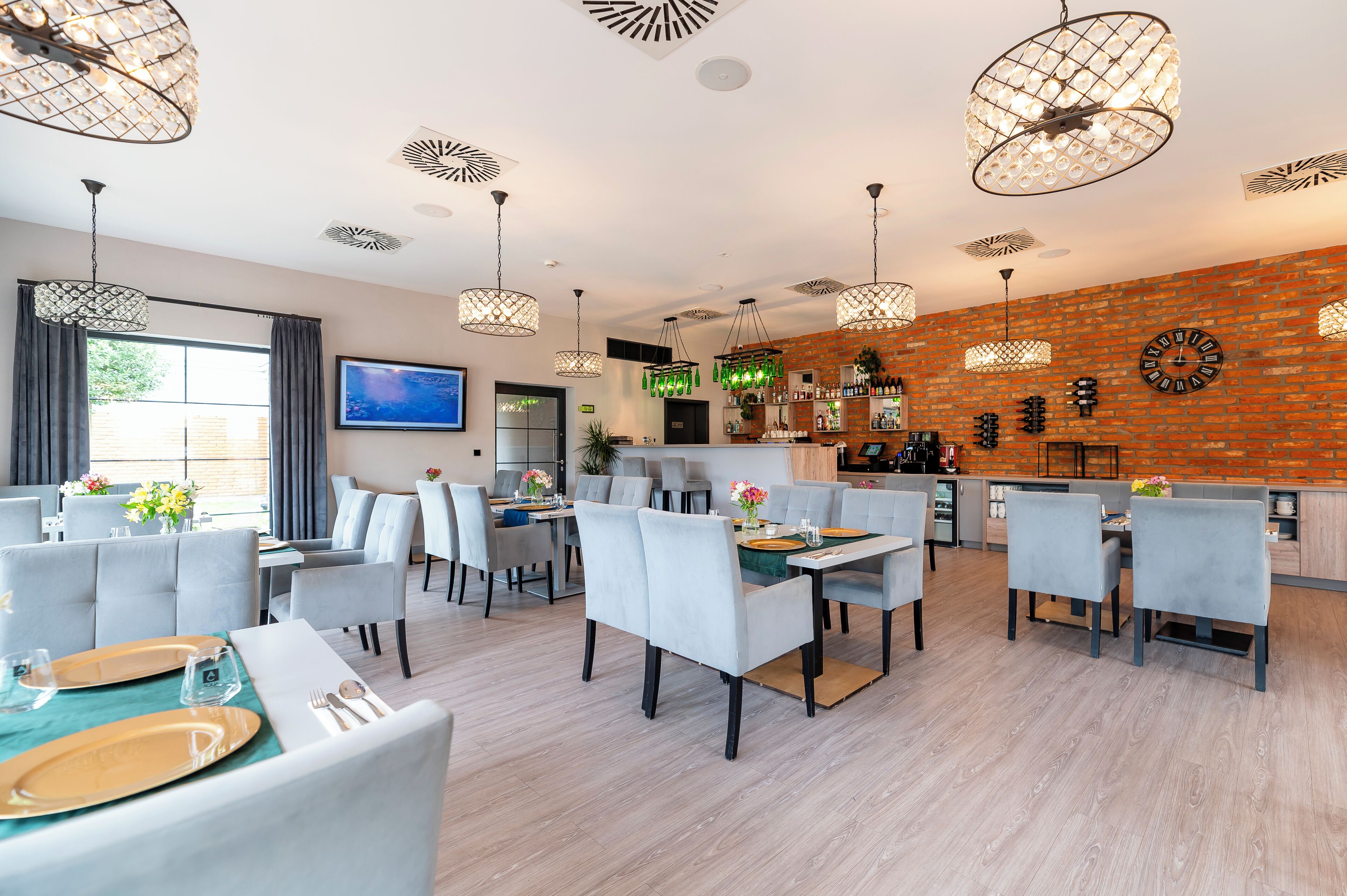 Uma sala de jantar moderna com cadeiras e mesas estofadas em cinza, uma parede de tijolos com um relógio e luminárias pendentes.
