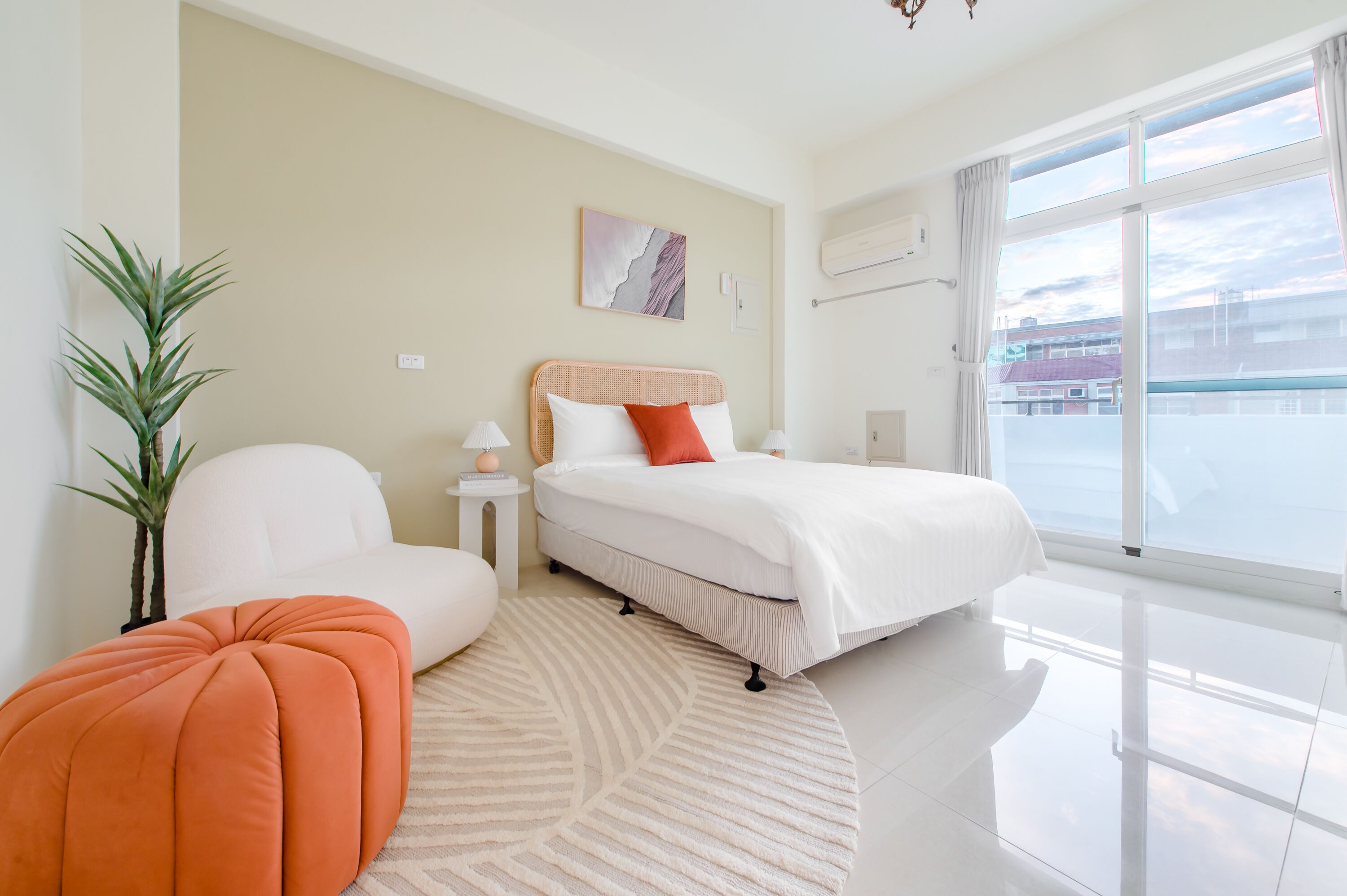 Quarto de hotel moderno com uma cama grande, uma poltrona branca, um banquinho laranja pequeno e uma janela ampla com vista para a cidade.