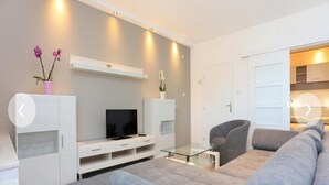 Uma sala de estar moderna com um sofá cinzento, uma televisão de tela plana e uma mesa de centro de vidro. Há armários brancos com portas de vidro e um vaso rosa com flores à esquerda.