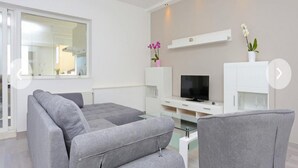 Uma sala de estar moderna com um sofá cinzento, uma mesa de centro de vidro, uma televisão de tela plana e armários brancos.