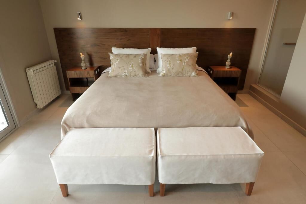Uma cama grande com duas mesas de cabeceira combinando, um painel de madeira na cabeceira e um banco aos pés da cama.