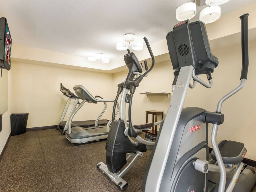 Sala de ginástica com esteiras e aparelhos elípticos, piso coberto de carpete e uma pequena mesa.