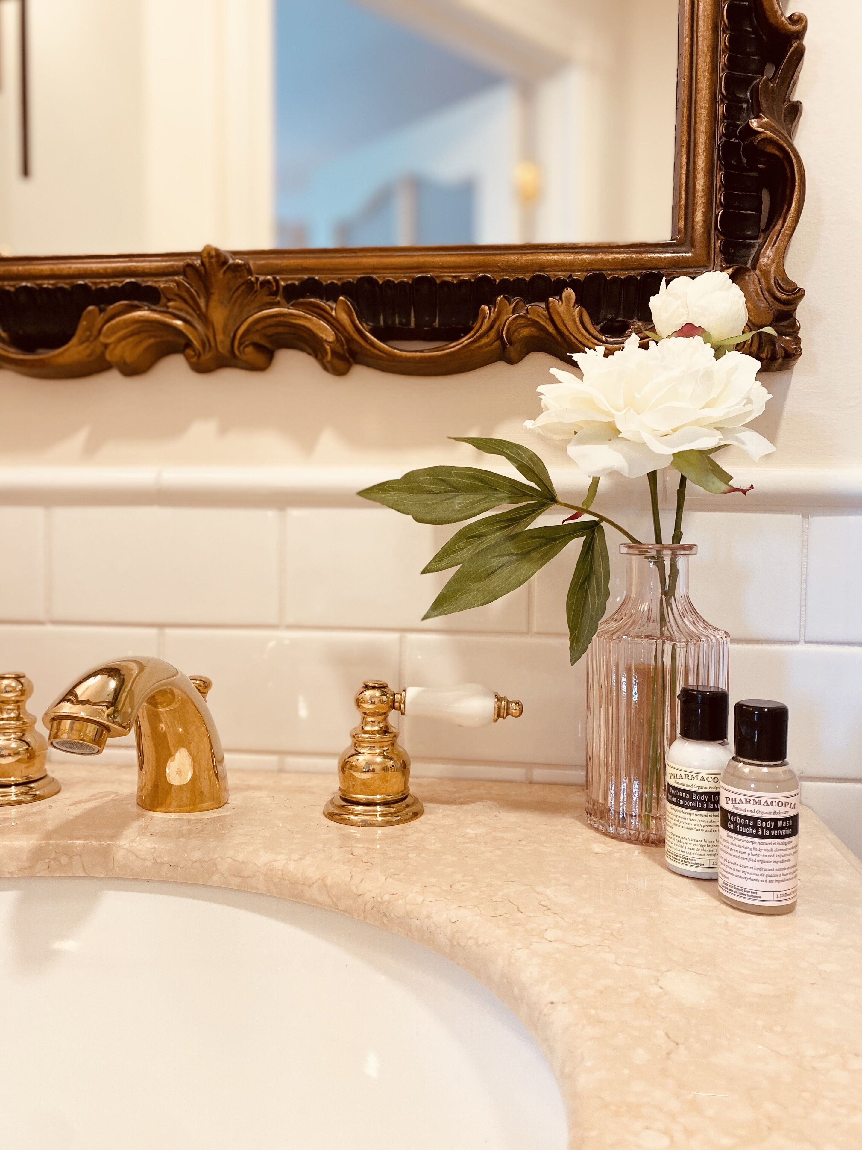 Um lavatório de banheiro com torneira dourada, pia branca e espelho com moldura ornamentada. Há dois frascos de gel de banho e um vaso com flores brancas no balcão.