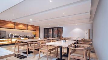 Uma área de refeições moderna com mesas e cadeiras de madeira, uma cozinha ao fundo e uma janela grande.