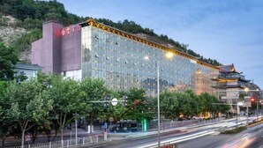 Um hotel moderno com fachada de vidro, situado num cruzamento de rua com semáforos e árvores.