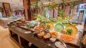Buffet com diversos pratos e utensílios, cercado por plantas decorativas e uma vitrine de vidro.