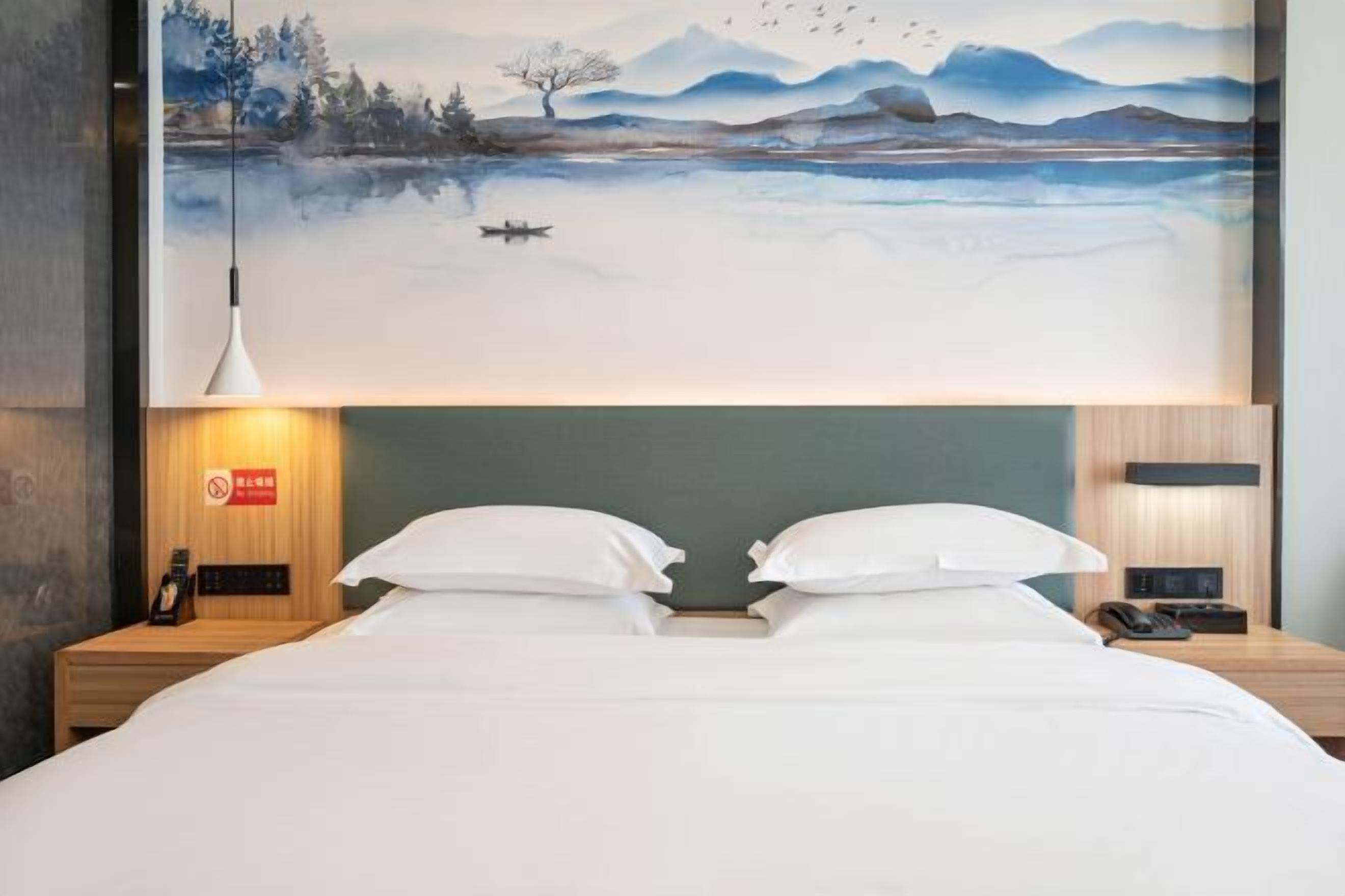 Quarto de hotel com uma cama grande, duas mesas de cabeceira, um criado-mudo, uma luminária na parede e um quadro de uma paisagem.